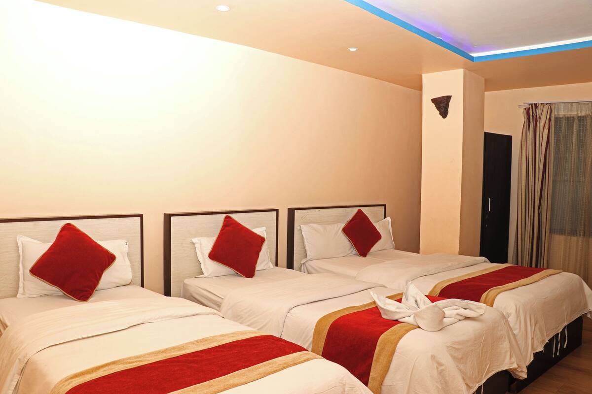Quarto de hotel com três camas, cada uma com roupa de cama branca e detalhes vermelhos, e um espelho grande acima das camas.