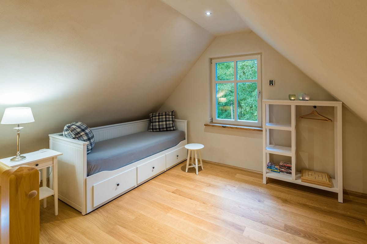 Um quarto de sótão acolhedor com cama embutida, uma pequena mesa com abajur, uma janela com vista para o verde e uma estante branca.