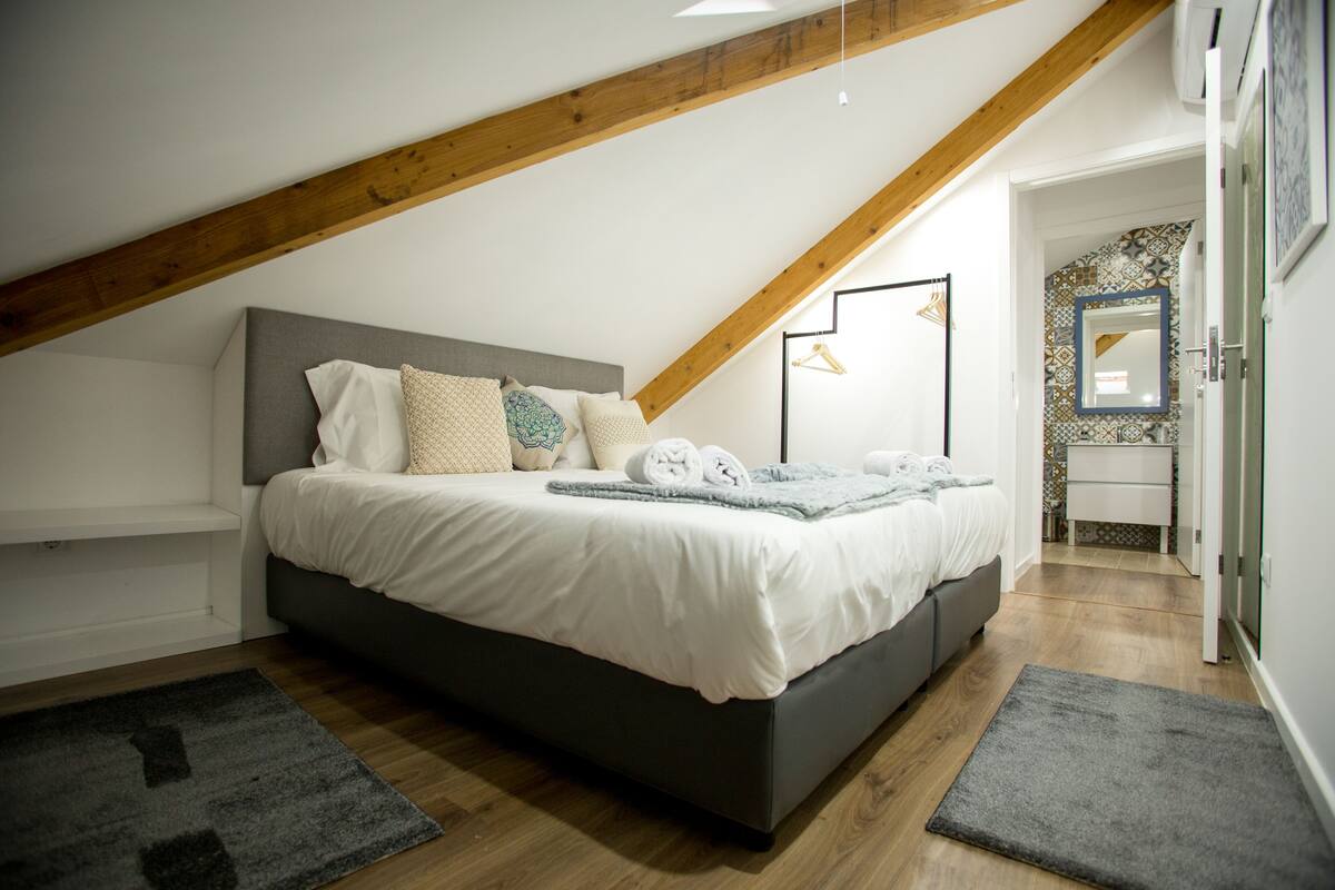 Quarto com teto inclinado, cama com roupa de cama branca e chão de madeira.