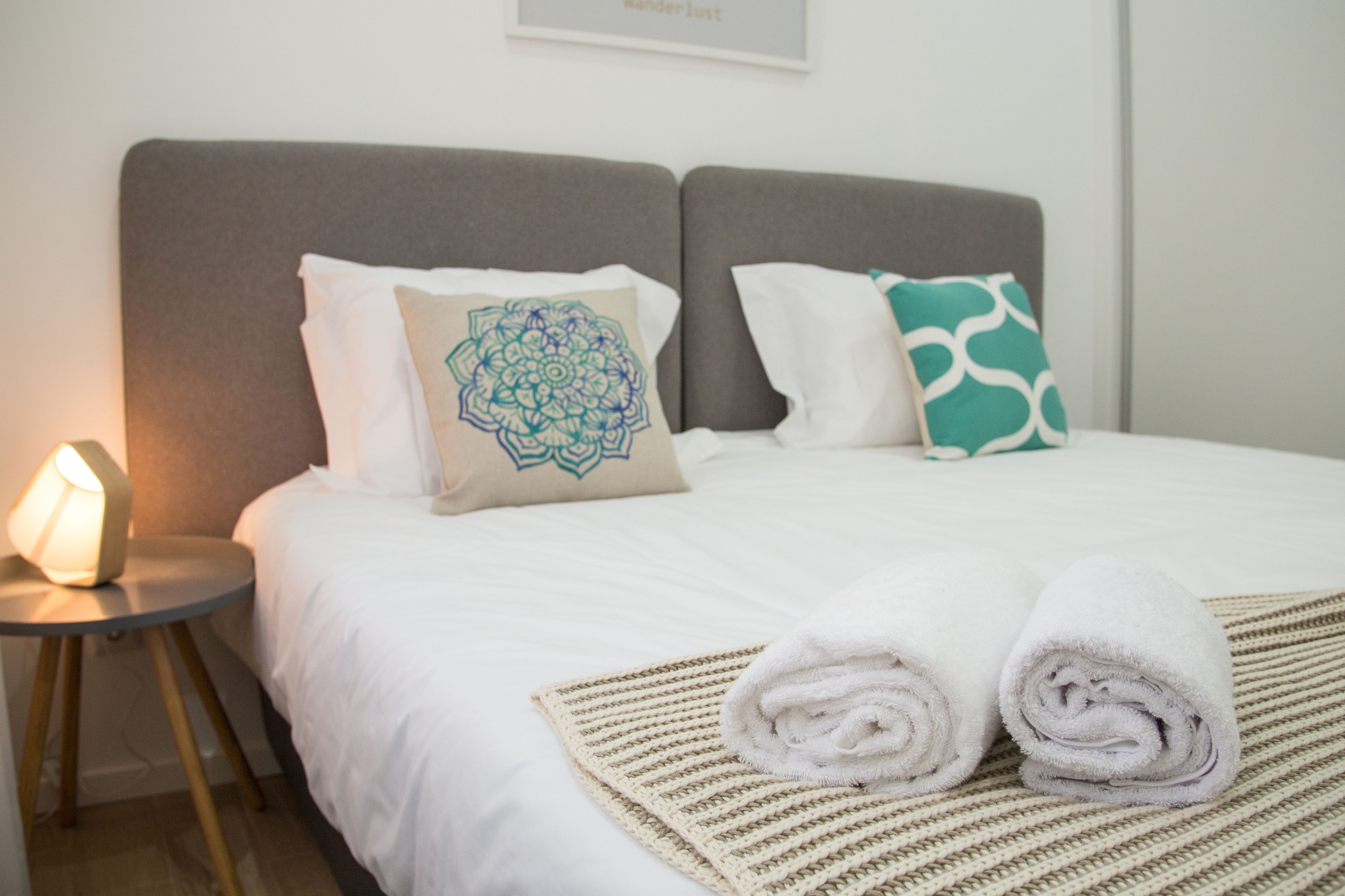 Cama bem arrumada com almofadas decorativas, abajur e toalhas enroladas sobre um tapete tecido.