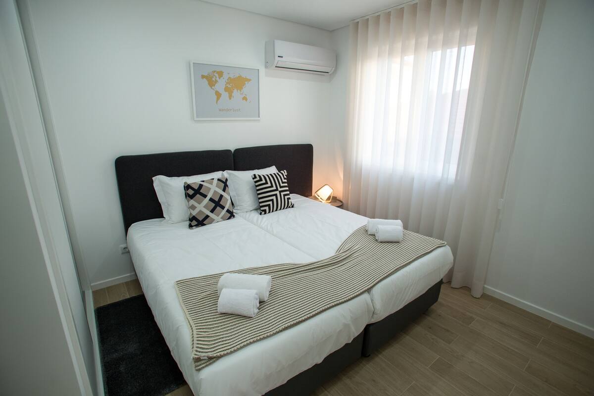 Uma cama bem arrumada com um mapa-múndi na parede, um ar condicionado e uma janela com cortinas.