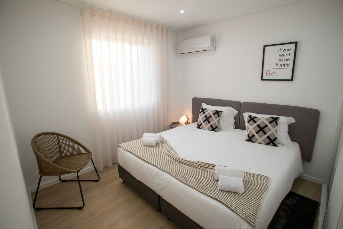 Quarto de hotel bem arrumado com uma cama, uma cadeira, mesa de cabeceira com abajur e ar-condicionado fixado na parede.