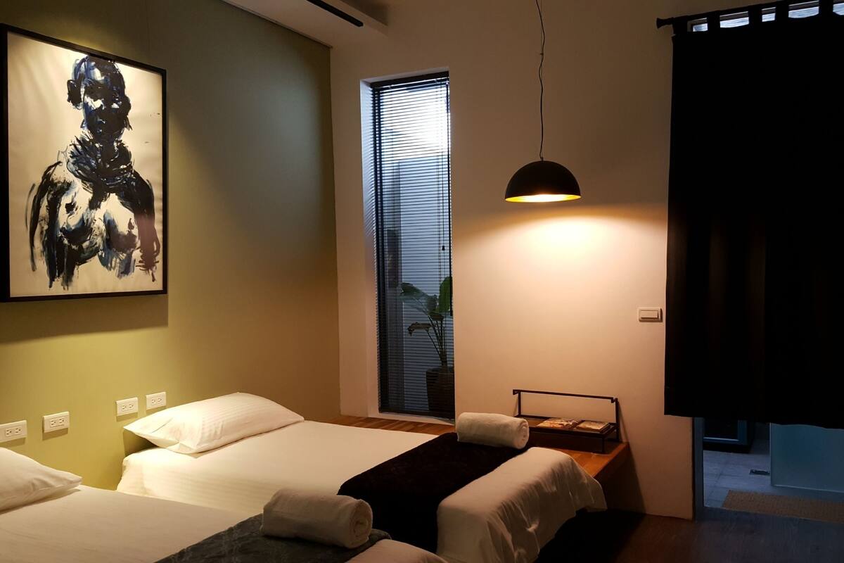 Quarto de hotel com duas camas, um quadro abstrato grande, uma janela com persianas, uma mesa de cabeceira com abajur e vista para o exterior.