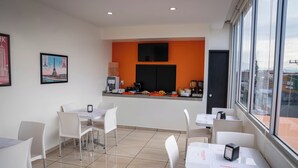 Uma área de refeições moderna, com cadeiras e mesas brancas, bancada de cozinha com eletrodomésticos e vista através de grandes janelas.