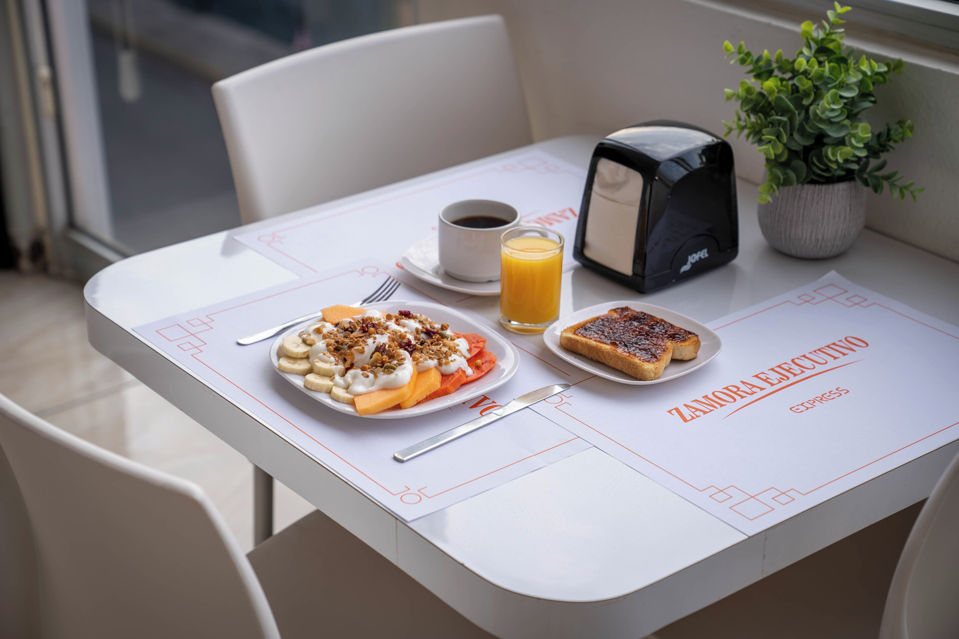 Uma mesa de café da manhã com um prato de comida, uma xícara de café, um copo de suco de laranja, uma torradeira e um vaso de planta.