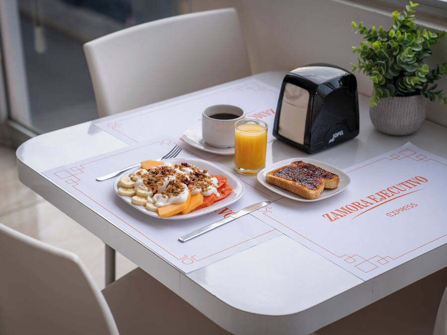 Uma mesa de café da manhã com um prato de comida, uma xícara de café, um copo de suco de laranja, uma torradeira e um vaso de planta.