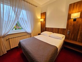 Quarto de hotel com cama, janela com cortinas leves, luminária na parede e radiador.