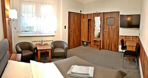 Quarto de hotel com cama, duas poltronas, uma mesa pequena, escrivaninha com cadeira, televisão e guarda-roupa.