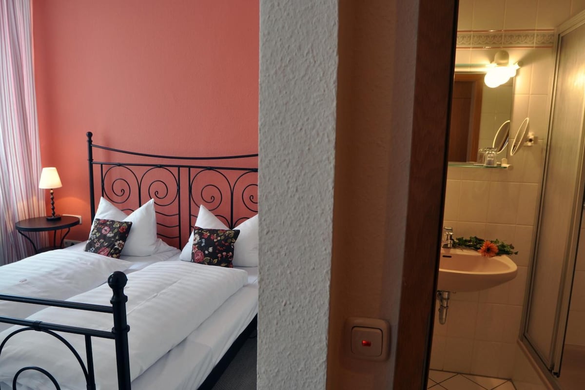 Uma cama com estrutura metálica e roupa de cama branca, uma pequena mesa de cabeceira com abajur, e uma parede com padrão decorativo.