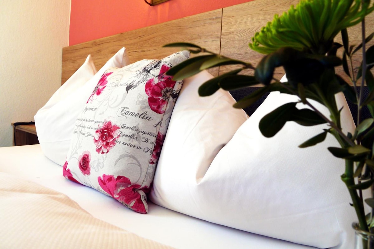 Cama com roupa de cama branca e duas almofadas, uma com estampas e texto florais, e um vaso de planta ao lado.