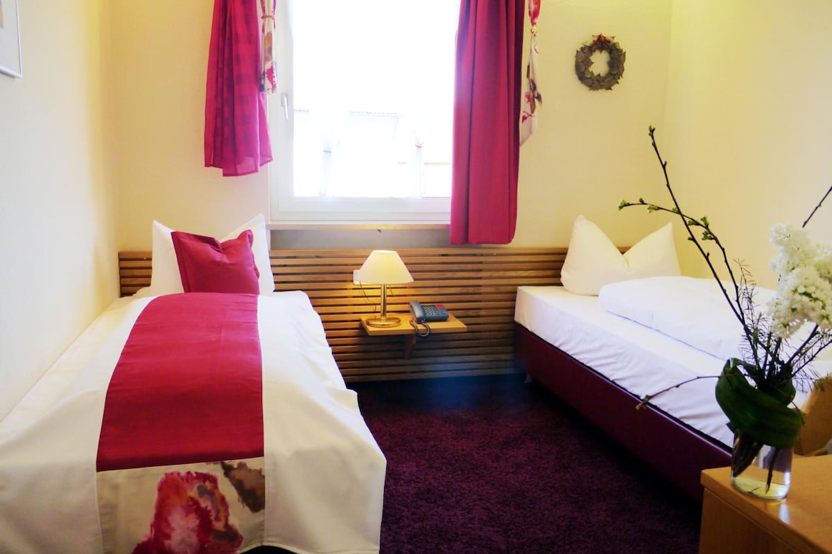 Quarto de hotel com duas camas, cabeceira de madeira, mesa de cabeceira pequena com abajur, telefone e um vaso com flores.