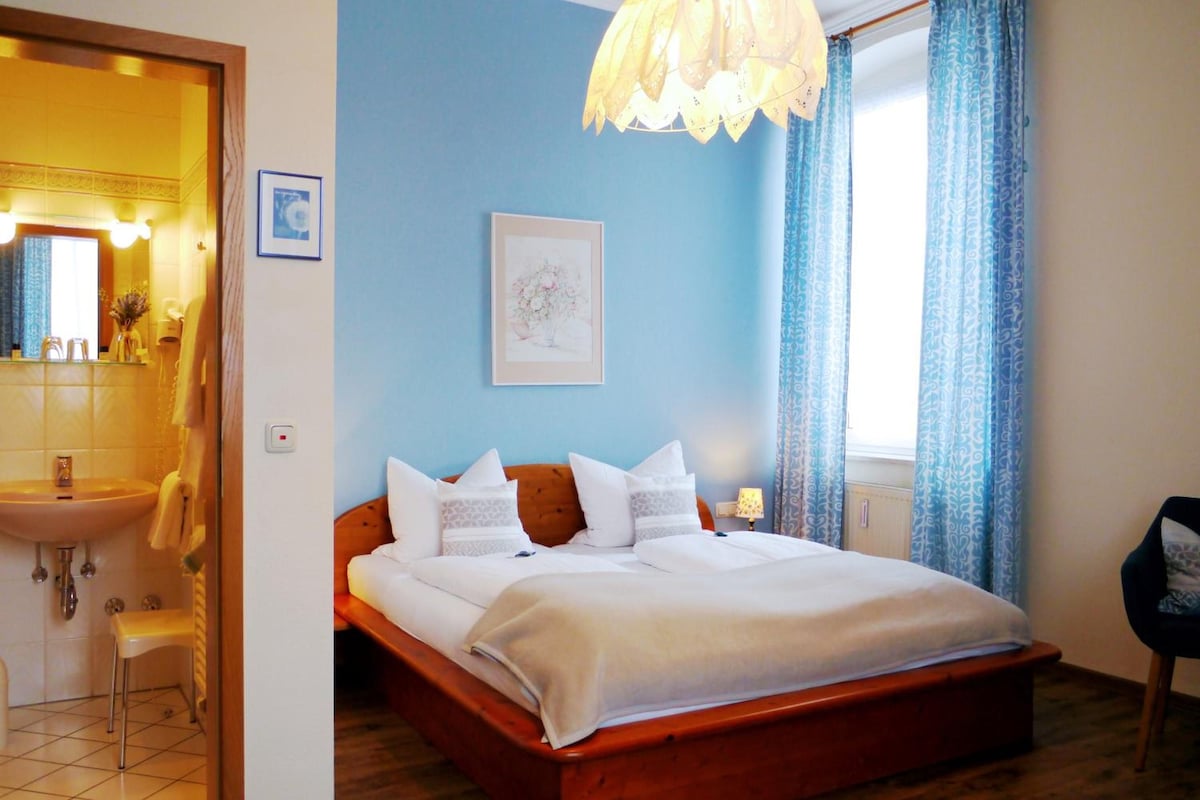 Quarto com cama de madeira, roupa de cama branca, mesa de cabeceira, luminária e janela com cortinas.