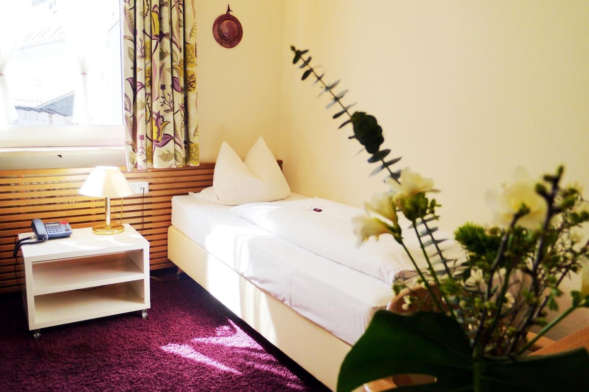 Quarto com cama, mesa de cabeceira com luminária, telefone e janela com cortinas.