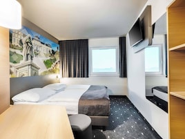 Quarto de hotel com duas camas, uma escrivaninha, uma televisão e um quadro de um edifício.