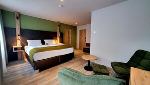 Quarto de hotel moderno com uma cama grande, uma mesinha de cabeceira, uma poltrona verde e uma pequena mesa redonda.