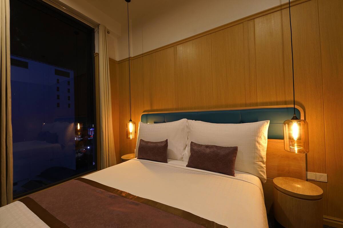 Quarto de hotel com uma cama grande, cabeceira de madeira, duas luminárias laterais e vista da cidade por uma janela ampla.