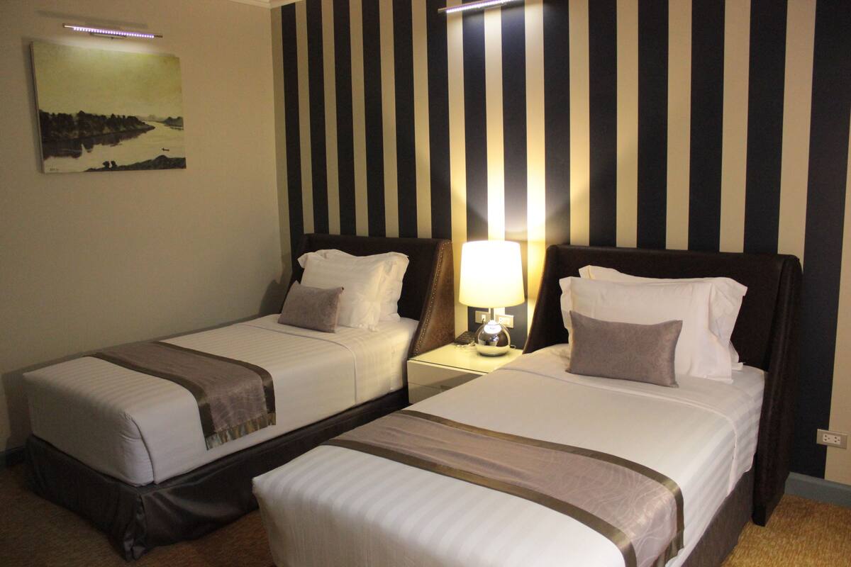 Quarto de hotel com duas camas de solteiro, mesa de cabeceira com abajur, pintura na parede e papel de parede com padrão listrado.