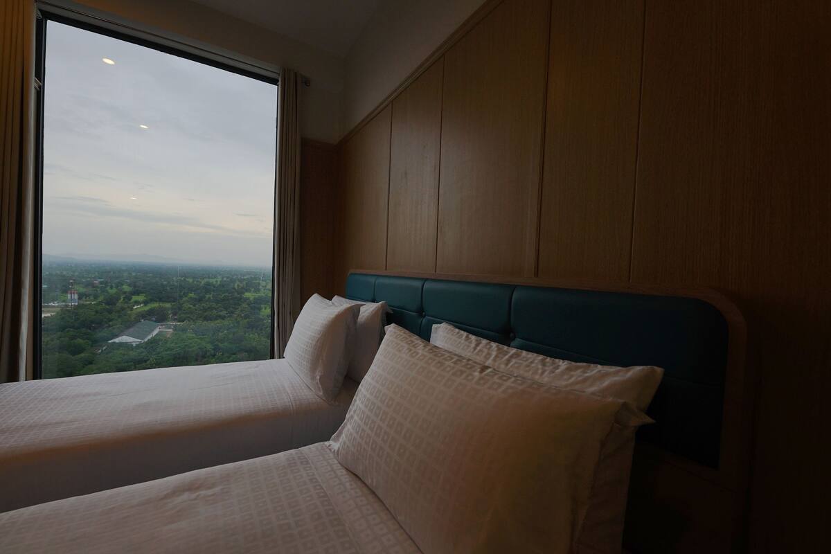 Quarto de hotel com uma janela grande que oferece vista para a cidade, uma cama com cabeceira verde-água e uma parede de painéis de madeira.