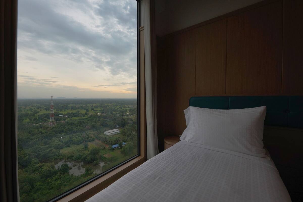 Quarto de hotel com uma janela grande que oferece vista para uma paisagem com vegetação e uma torre distante. O quarto tem uma cabeceira de madeira e uma cama bem arrumada com roupa de cama branca.