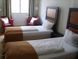 Quarto de hotel com duas camas de solteiro, uma mesa de cabeceira, uma cadeira vermelha e janela com cortinas.