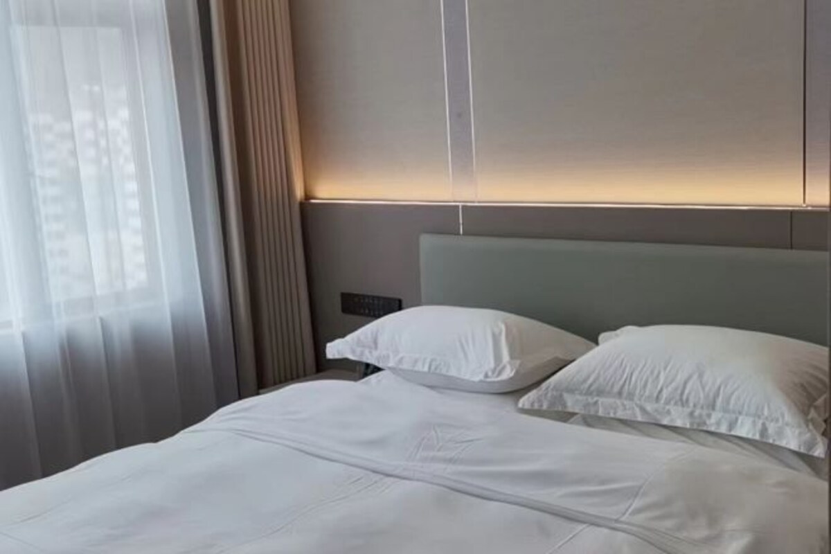 Quarto de hotel com cama, roupa de cama branca, cabeceira com luminária, mesa de cabeceira e janela com cortinas.