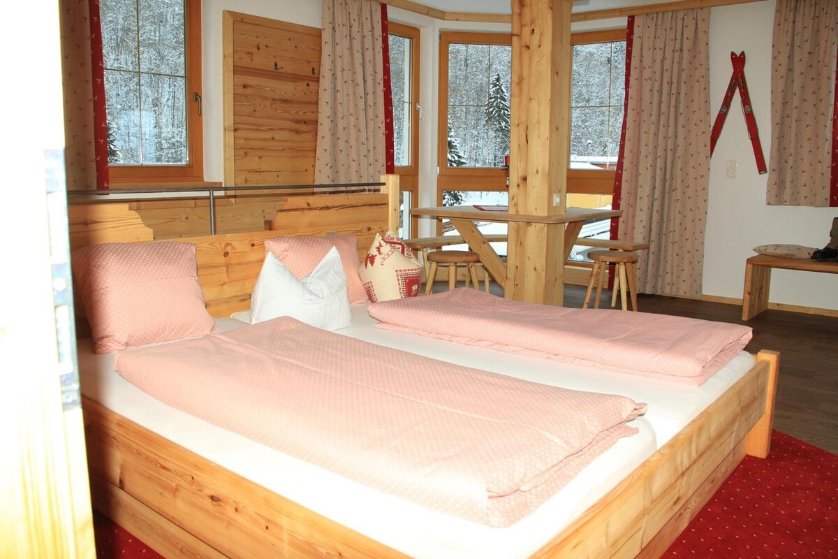 Uma cama de casal com cabeceira e peseira de madeira, uma mesinha e uma cadeira.