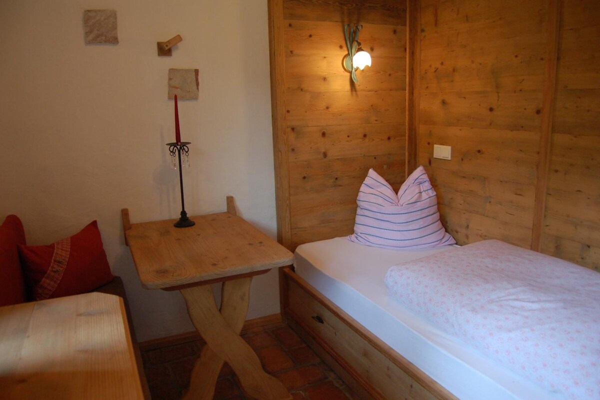 Quarto pequeno e acolhedor com cama de madeira, uma mesinha e luminária na parede.