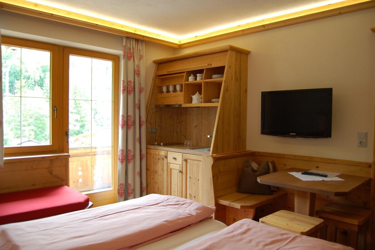 Quarto estilo cabana com cama, uma mesinha, televisão e janela com cortinas.