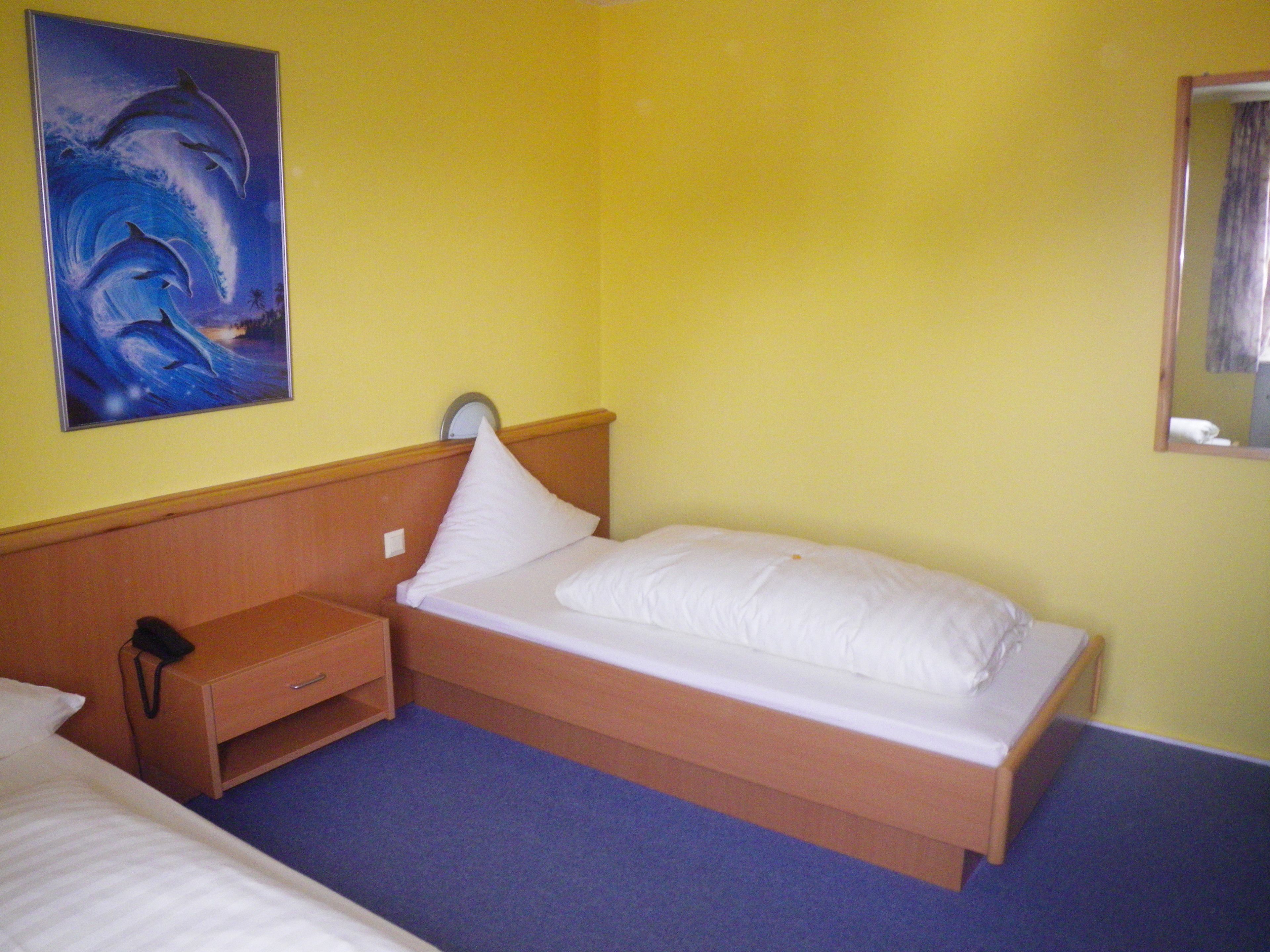 Uma cama de solteiro com cabeceira de madeira, uma mesa de cabeceira com abajur e um quadro de golfinhos na parede.