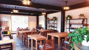 Restaurante com mesas e cadeiras de madeira, iluminação pendente e prateleiras com plantas e utensílios de cozinha.