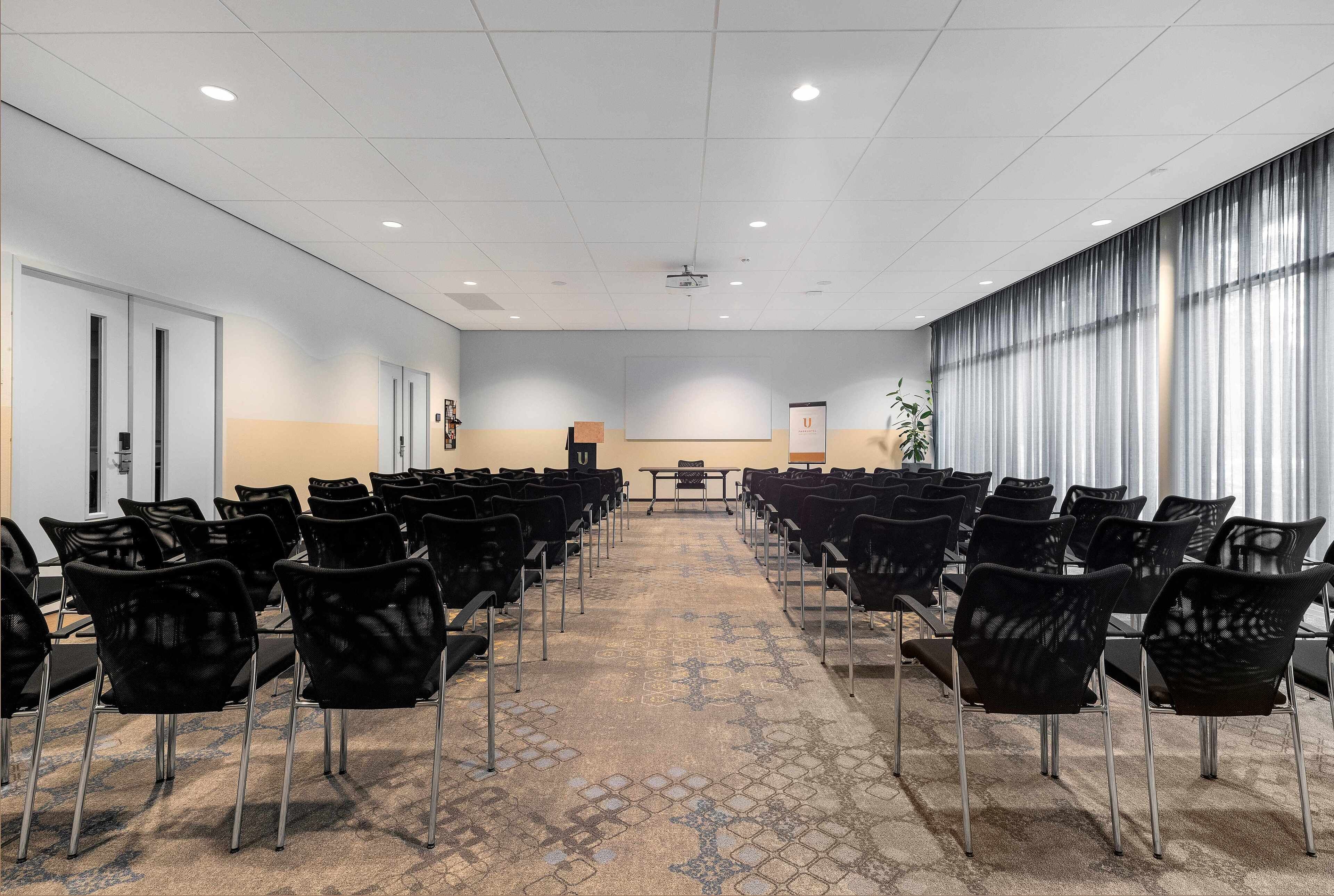 Uma sala de conferências com fileiras de cadeiras voltadas para um púlpito, piso coberto de carpete e uma janela grande de um lado.