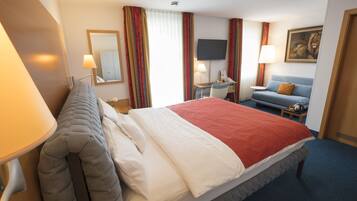 Quarto de hotel com uma cama grande, uma mesa, uma cadeira, um sofá, uma televisão e um quadro na parede.