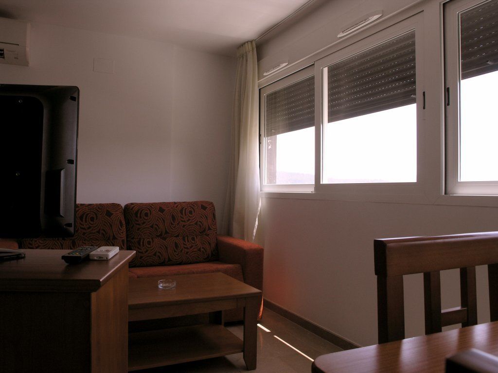Uma sala de estar com sofá, mesa de centro e televisão. Há duas janelas com persianas.