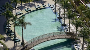 Vista aérea de um resort com piscina, espreguiçadeiras, palmeiras e uma ponte.