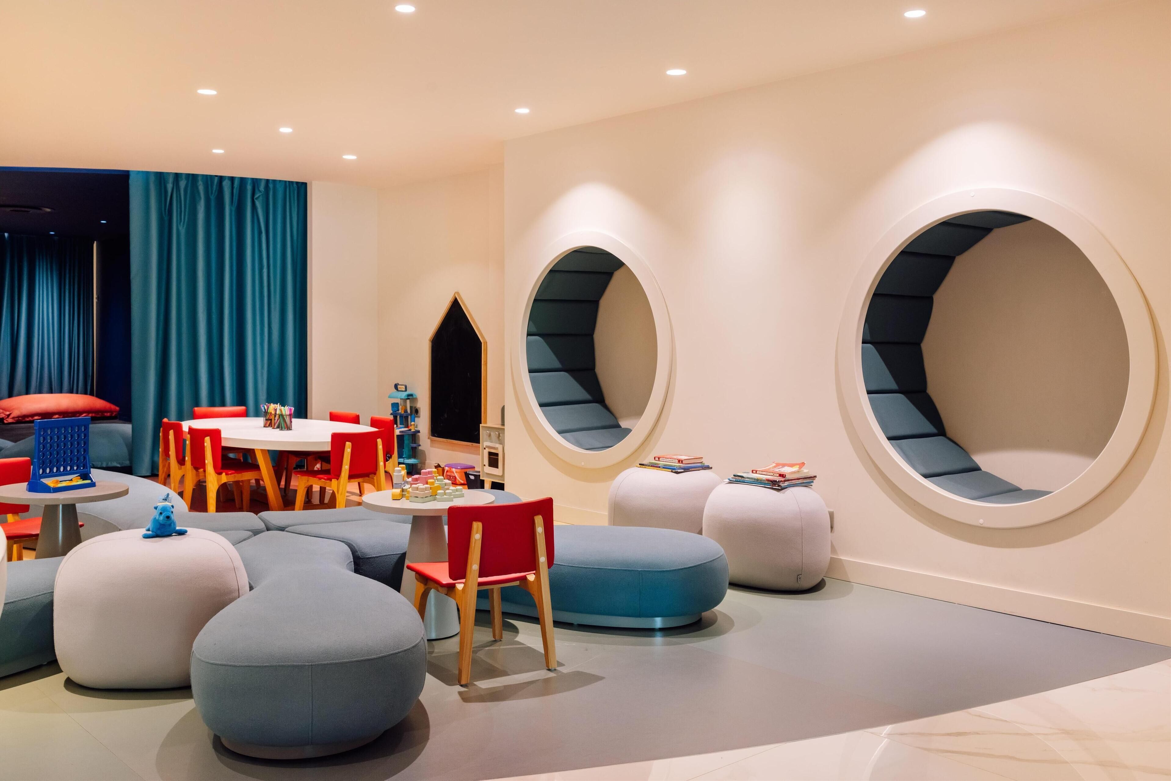 Uma sala de jogos infantil moderna com assentos redondos, uma mesa de brincar e uma grande janela com cortinas.