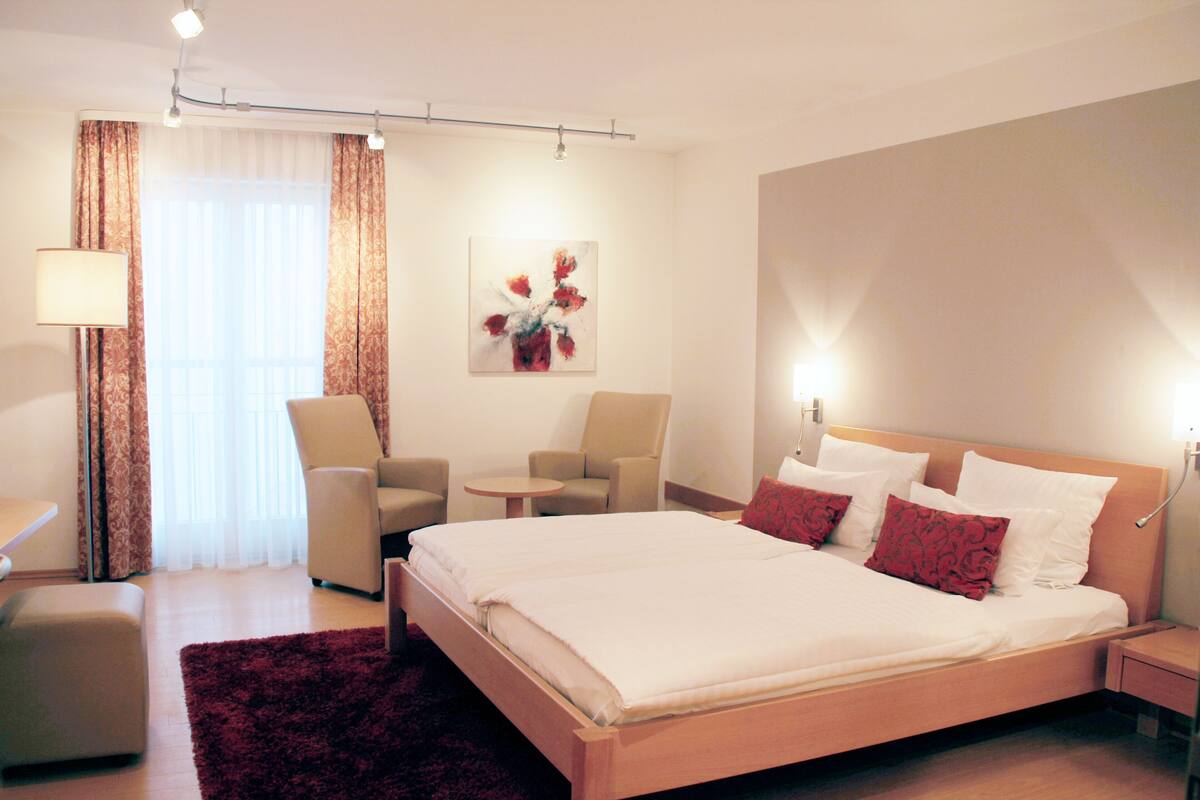 Quarto de hotel com uma cama, duas cadeiras, uma mesinha, um abajur e um quadro na parede.