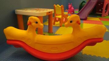 Uma área de recreação infantil com um barco amarelo, uma mesa pequena, cadeiras e um escorrega.