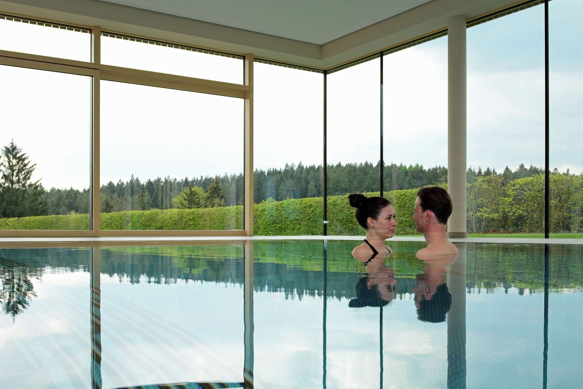 Um casal em uma piscina interna transparente, com janelas amplas e vista para uma paisagem arborizada.