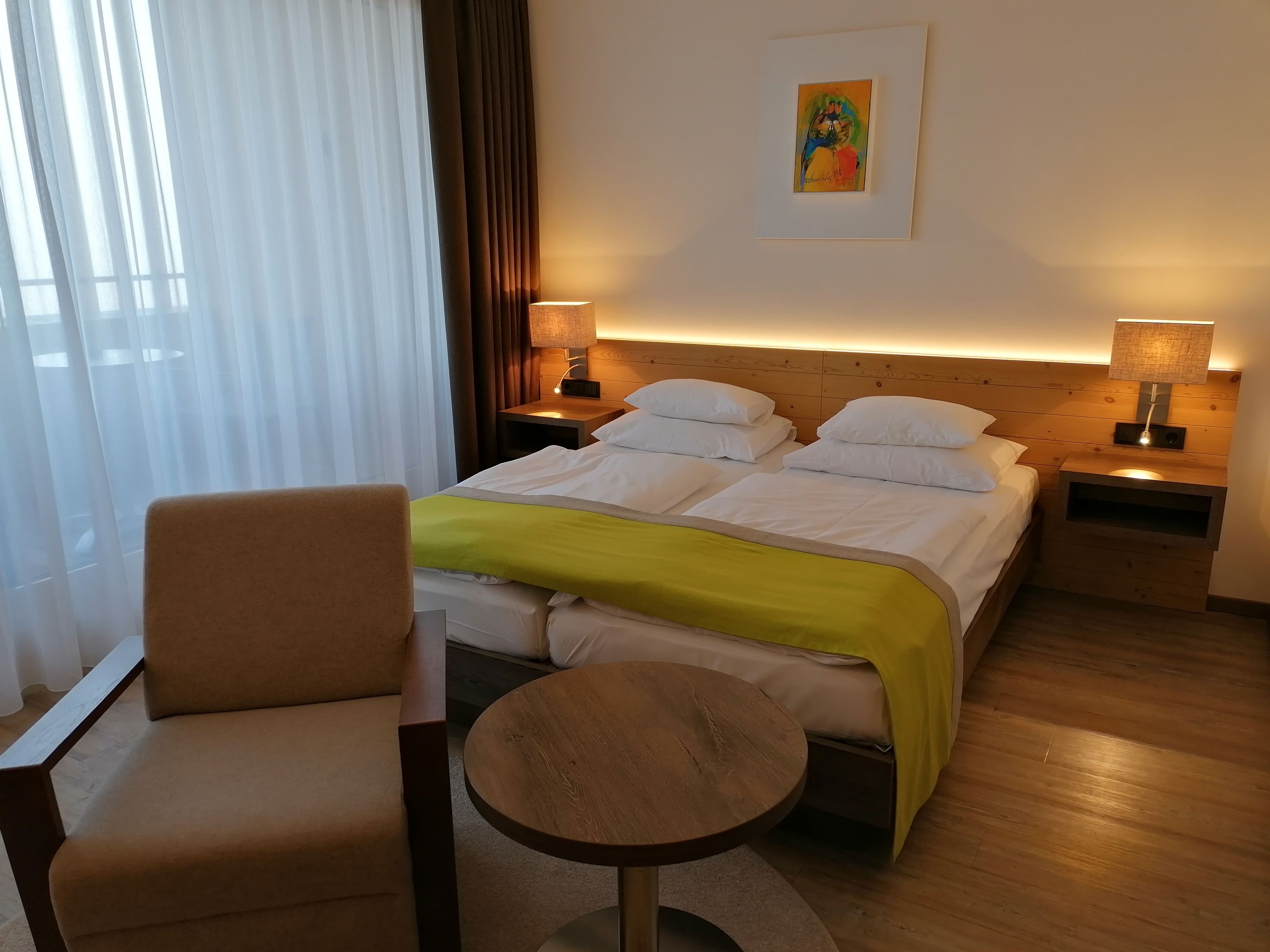 Quarto de hotel com cama, duas mesas de cabeceira com abajures, uma cadeira, uma mesinha redonda e um quadro na parede.