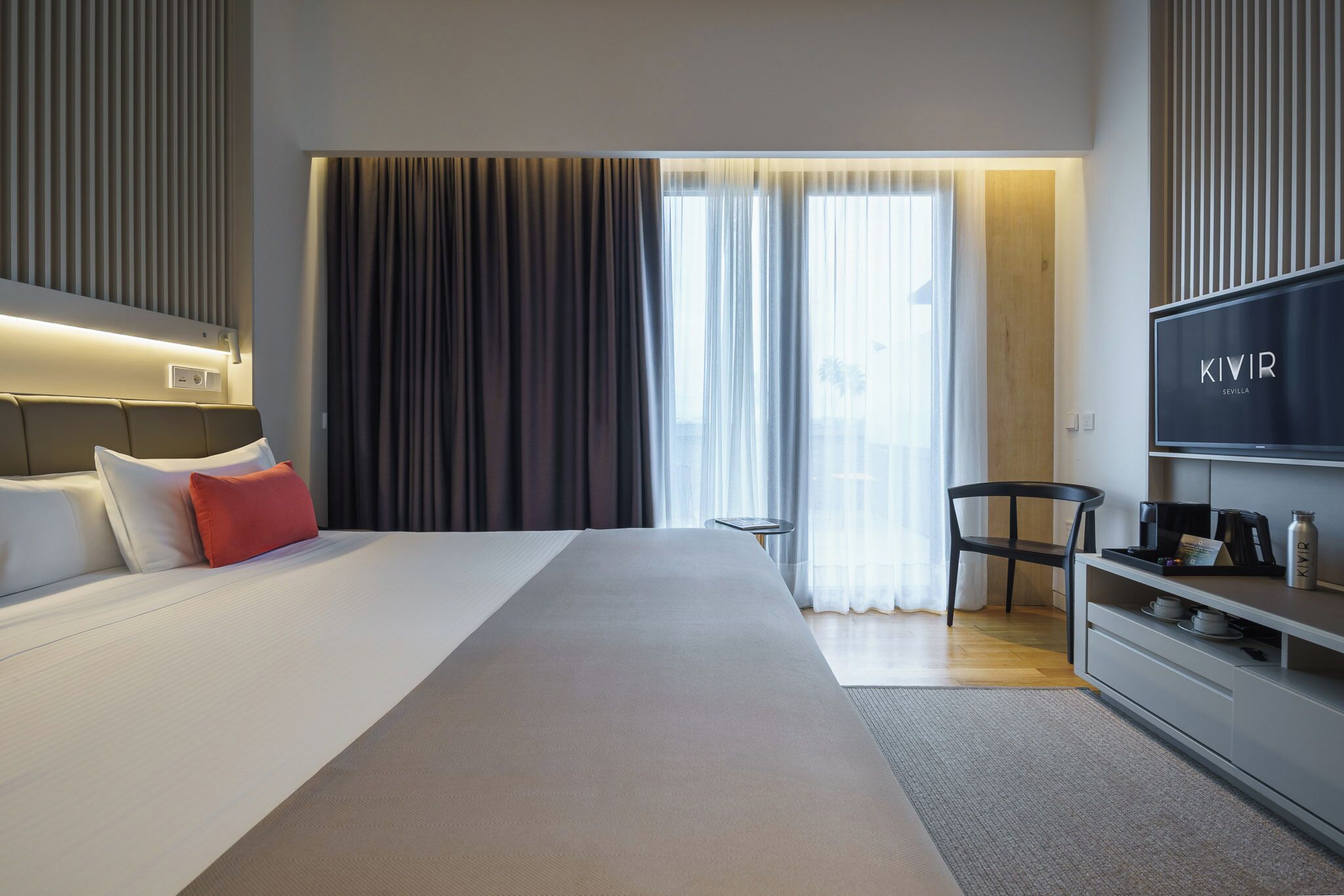 Quarto de hotel moderno com uma cama grande, TV, escrivaninha e vista para o exterior pela janela.