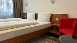 Quarto de hotel com cama, uma cadeira vermelha, uma mesinha e luminária fixada na parede.