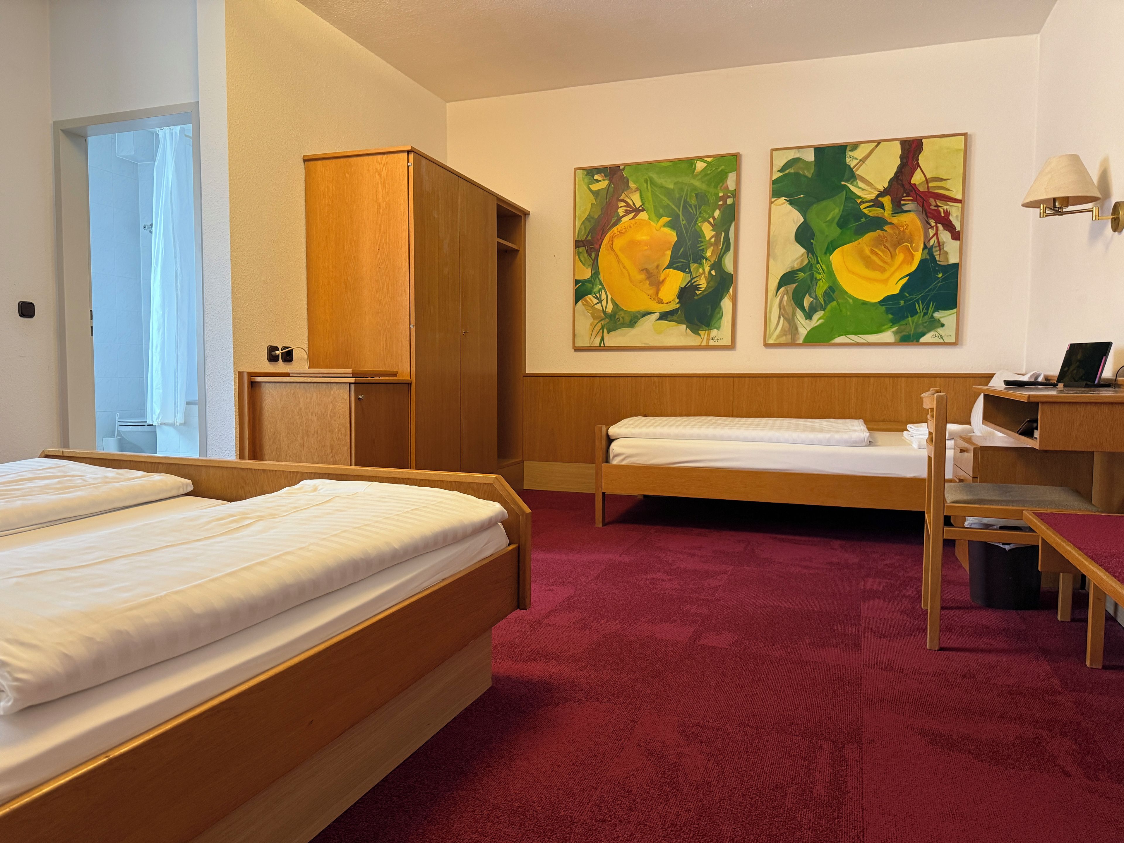 Quarto de hotel com duas camas, uma escrivaninha, uma cadeira, um guarda-roupa e duas pinturas nas paredes.