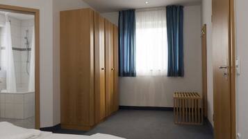 Quarto de hotel com chuveiro, guarda-roupa, janela com cortinas e uma pequena lixeira.