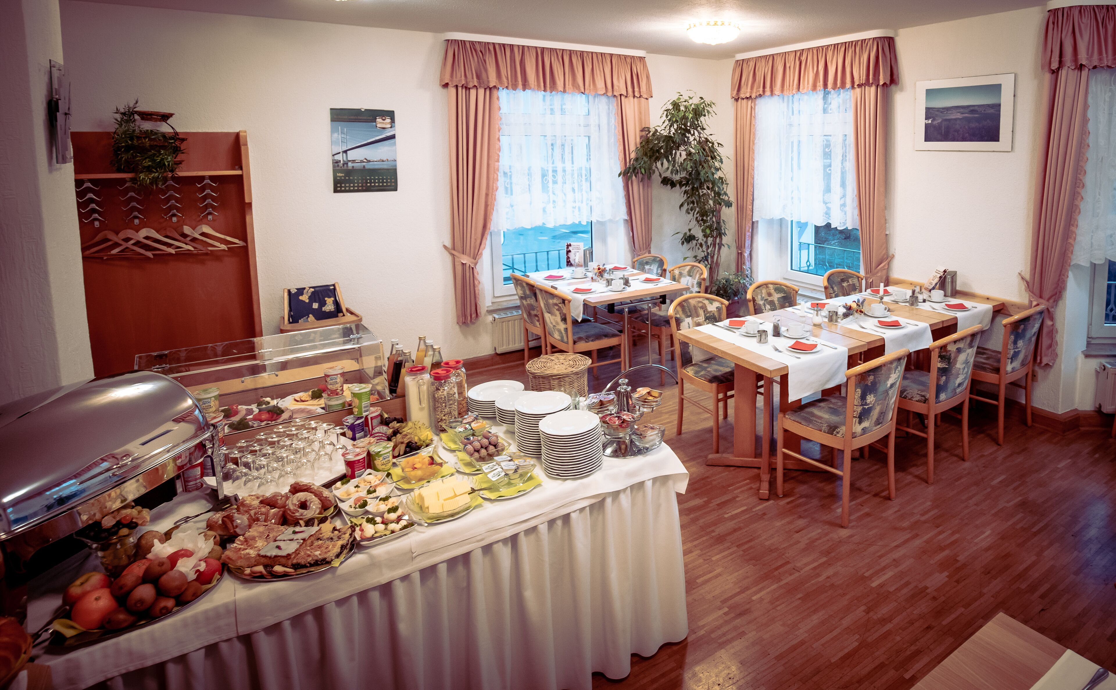 Uma área de refeições com mesa de buffet, mesas, cadeiras e grandes janelas com cortinas.