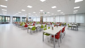 Uma sala de jantar ampla, com várias mesas e cadeiras, janelas grandes e um pilar central.