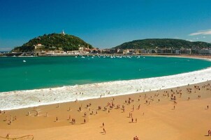 Uma praia com uma grande multidão, águas turquesas cristalinas e uma colina com edifícios ao fundo.