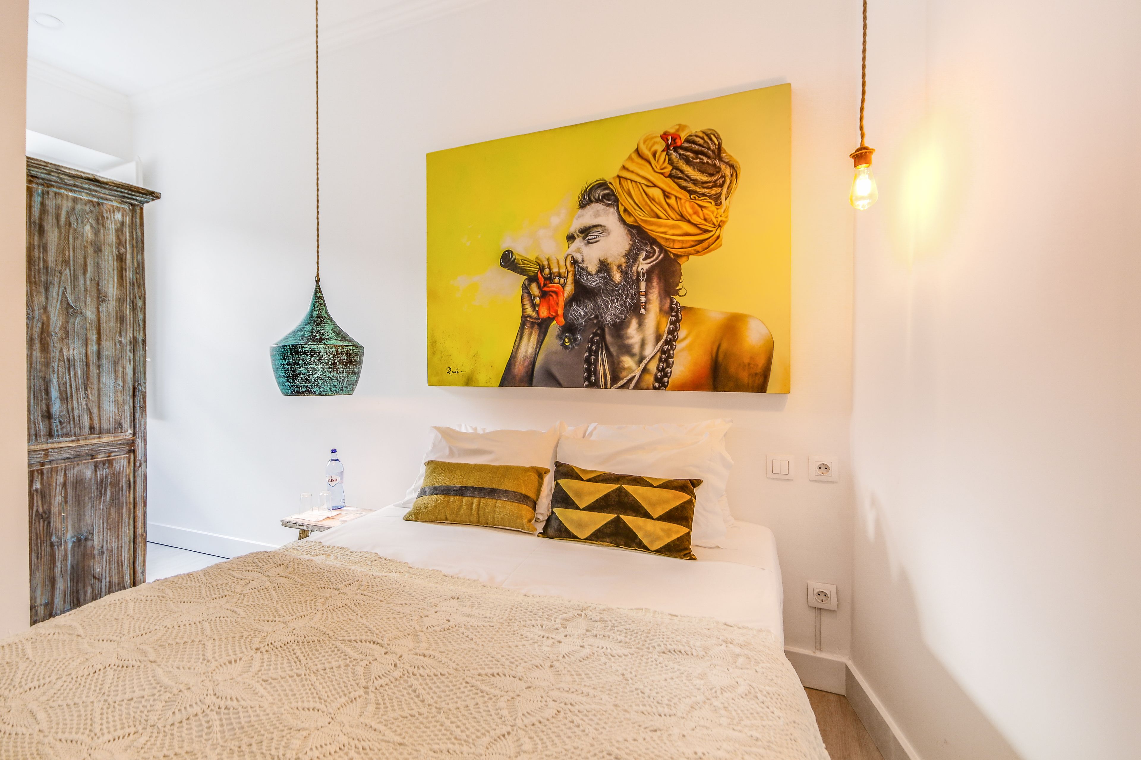 Quarto com cama, almofadas, pintura, luminária pendente e guarda-roupa de madeira.