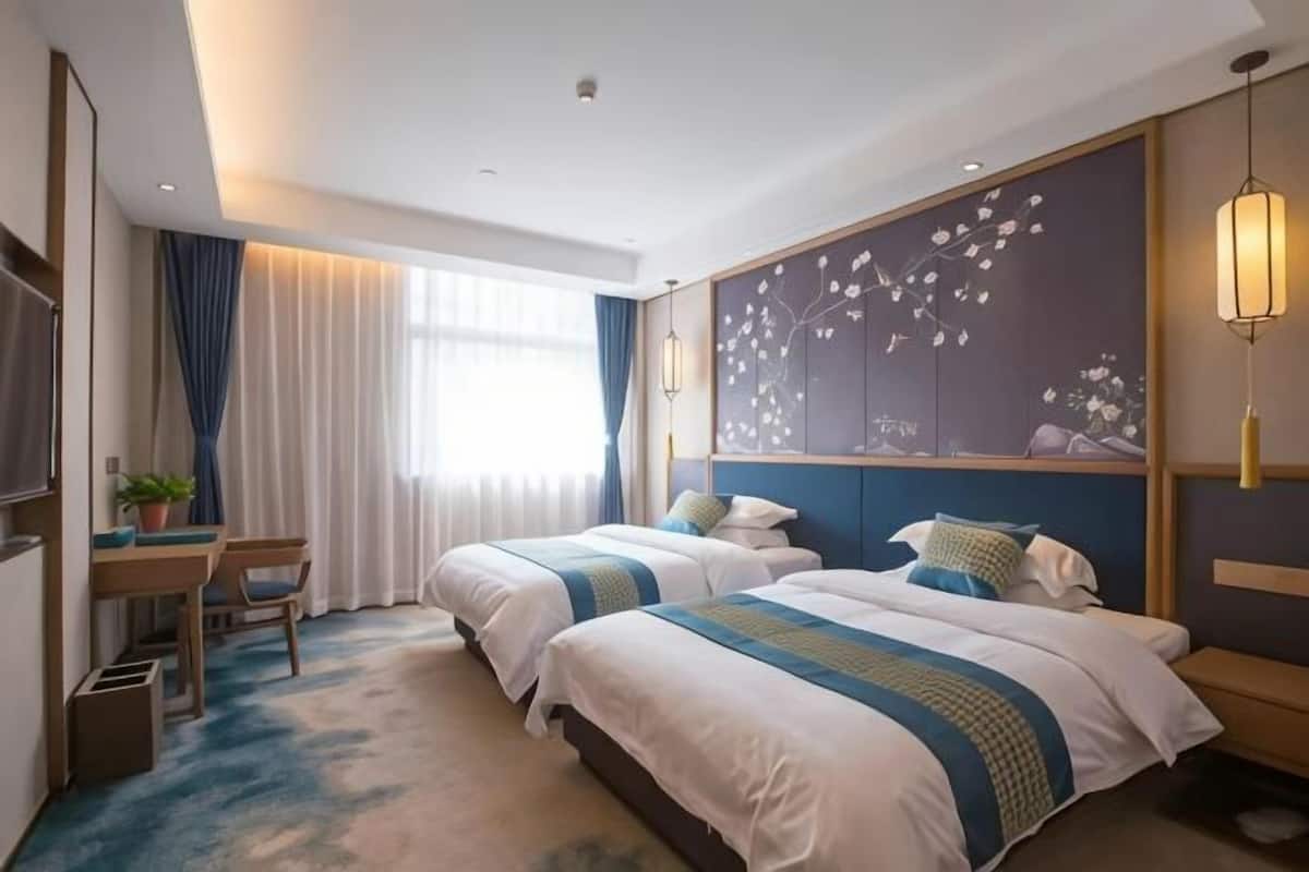 Quarto de hotel com duas camas, uma escrivaninha, uma cadeira, televisão e uma janela grande com cortinas.