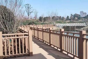Uma ponte de madeira com grades, que leva a um corpo d'água e uma vista distante de um parque com árvores e uma colina.