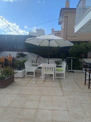 Um pátio externo com um guarda-sol branco, cadeiras brancas e uma mesa pequena.